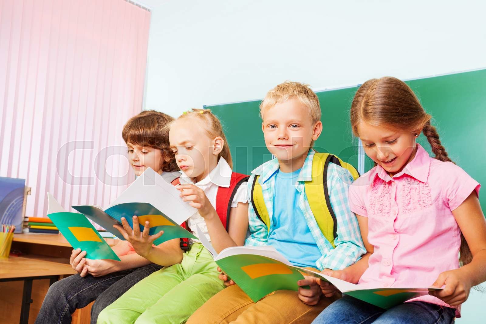 Four schoolchildren sit in row on desk and read | Stock image | Colourbox