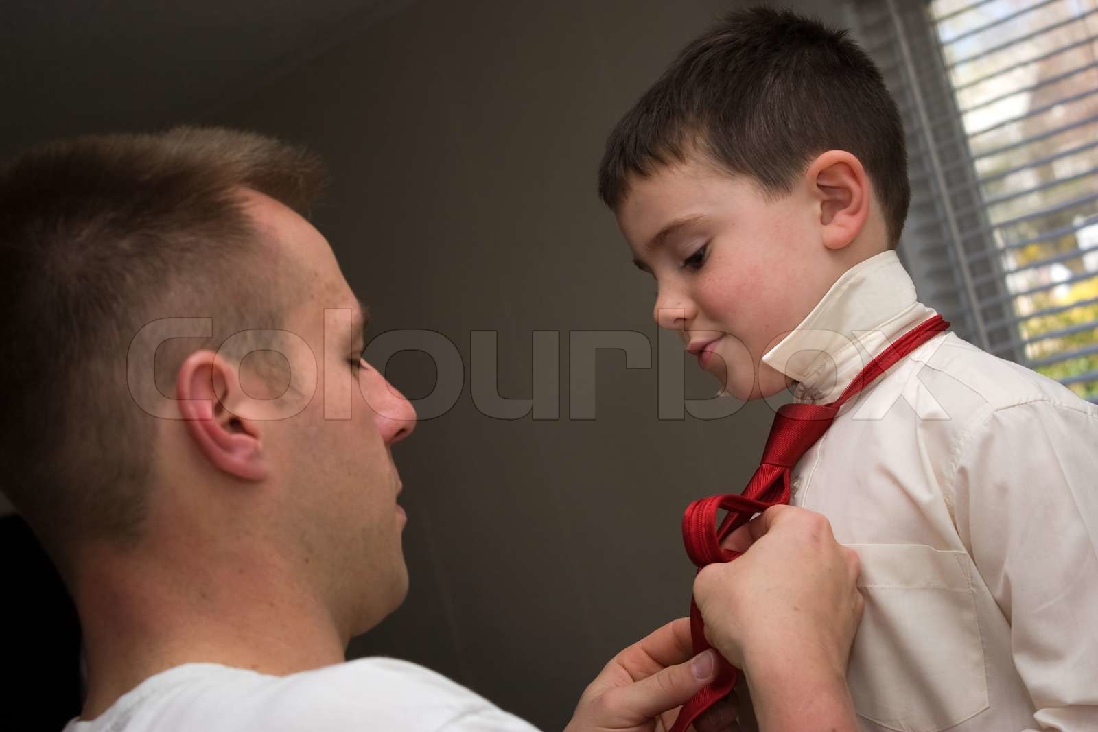 A young dad helps his son get ready by helping him tie his neck tie ...
