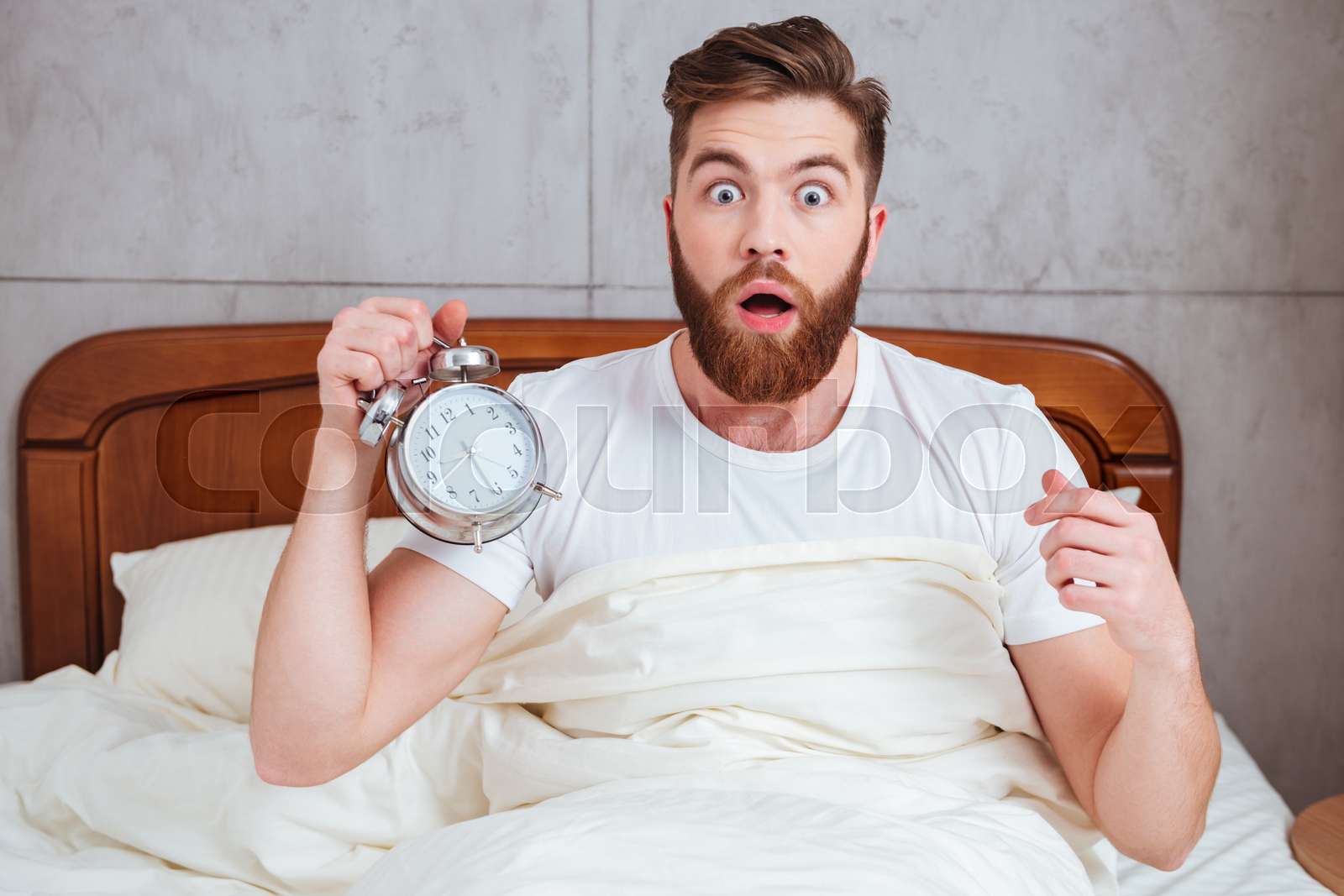 Confused man in bed holding alarm clock in hand | Stock image | Colourbox