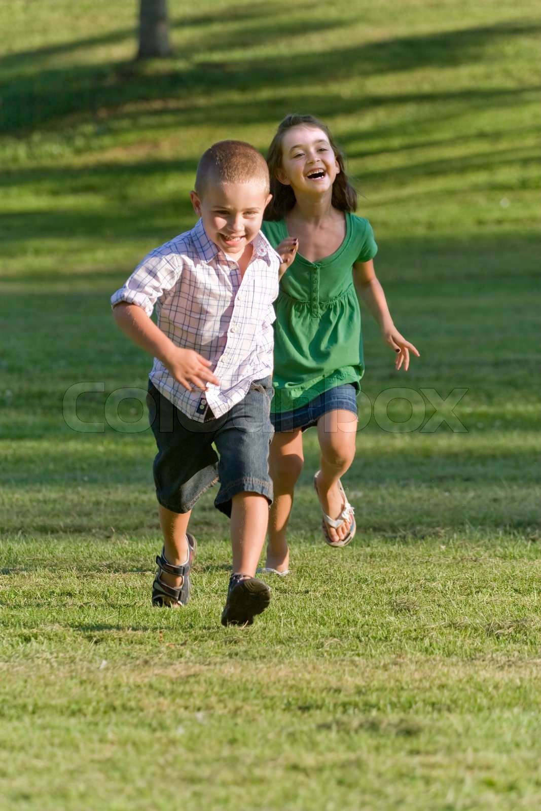 A young brother and sister running through a green grassy field with ...
