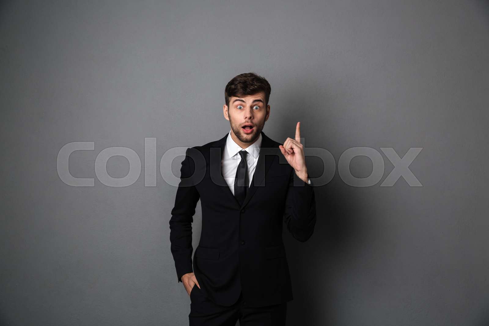 Amazed young man in suit pointing with finger upward, looking at camera ...