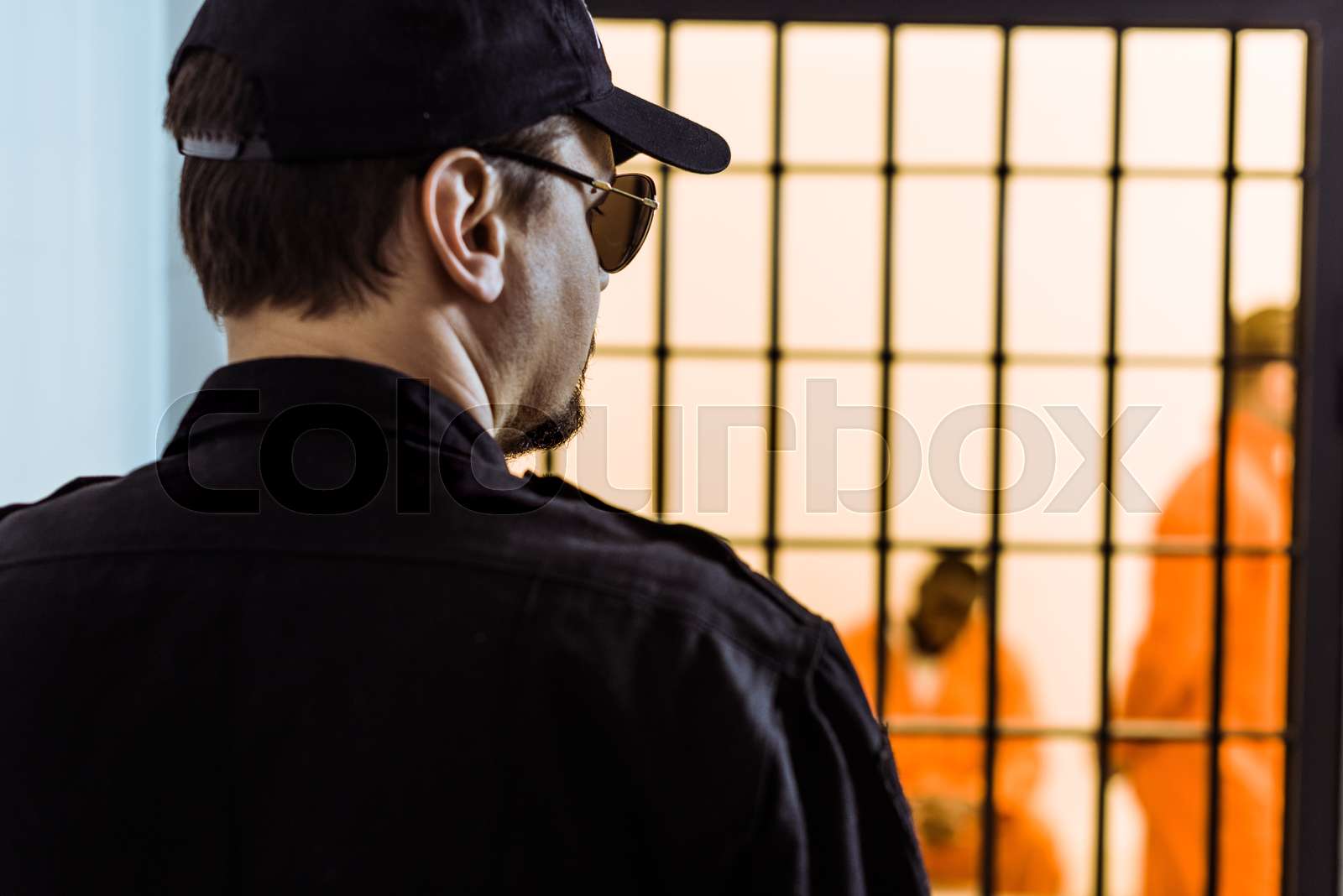 rear view of prison officer standing near prison cell with criminals ...