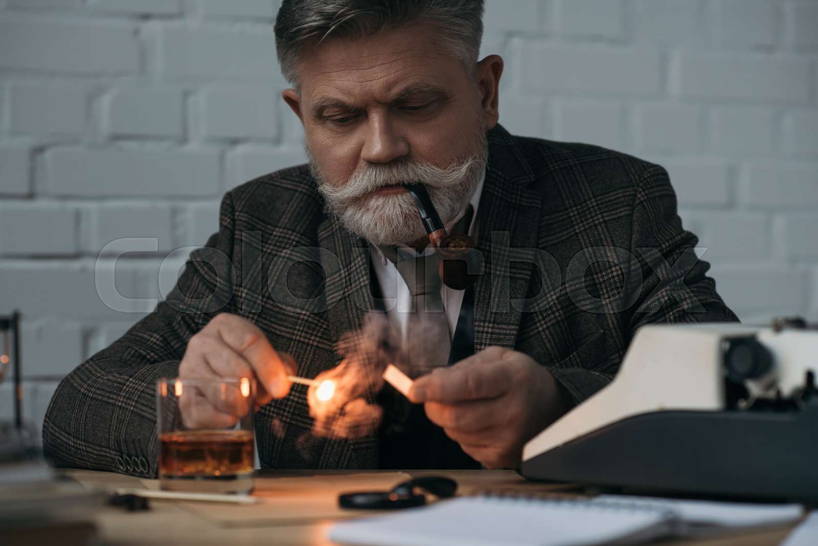 bearded senior writer with burning match smoking pipe at workplace ...