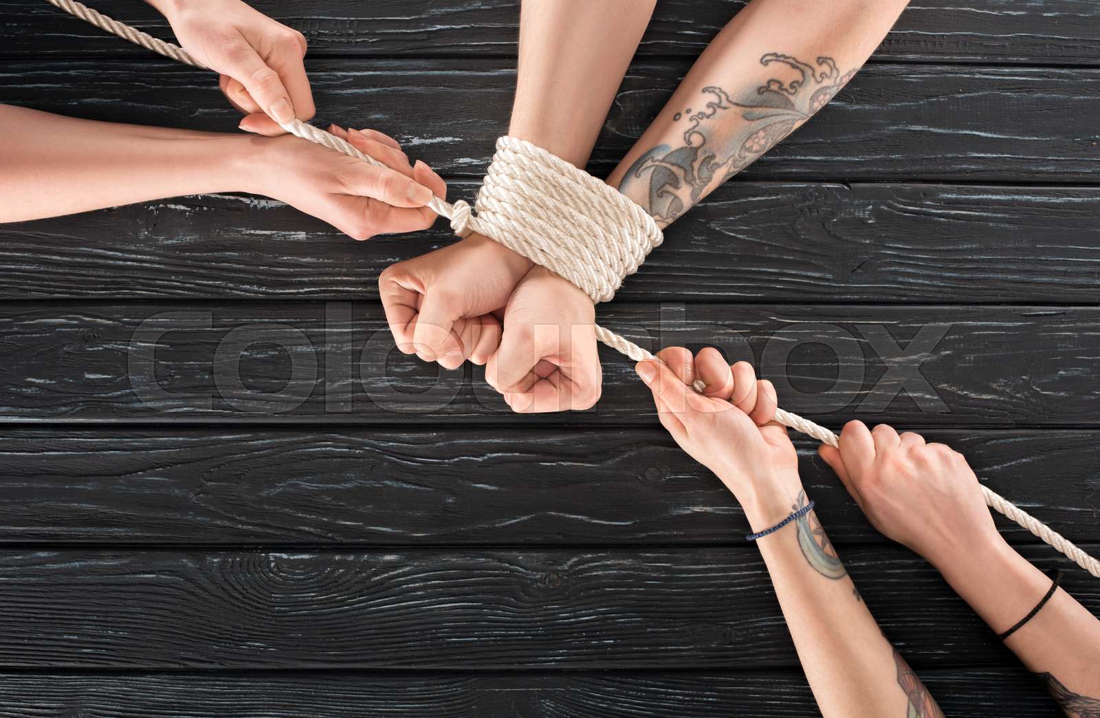 partial view of women tying rope around males hands on dark wooden ...
