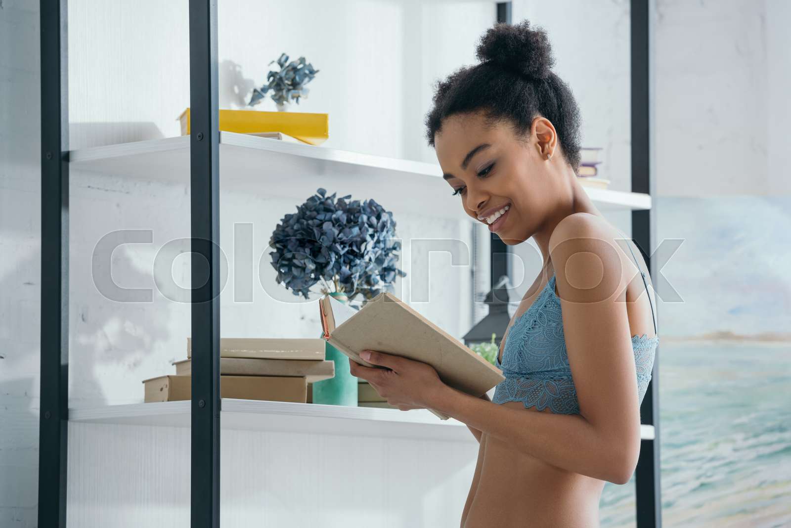 smiling african american girl in underwear reading book near bookshelf ...