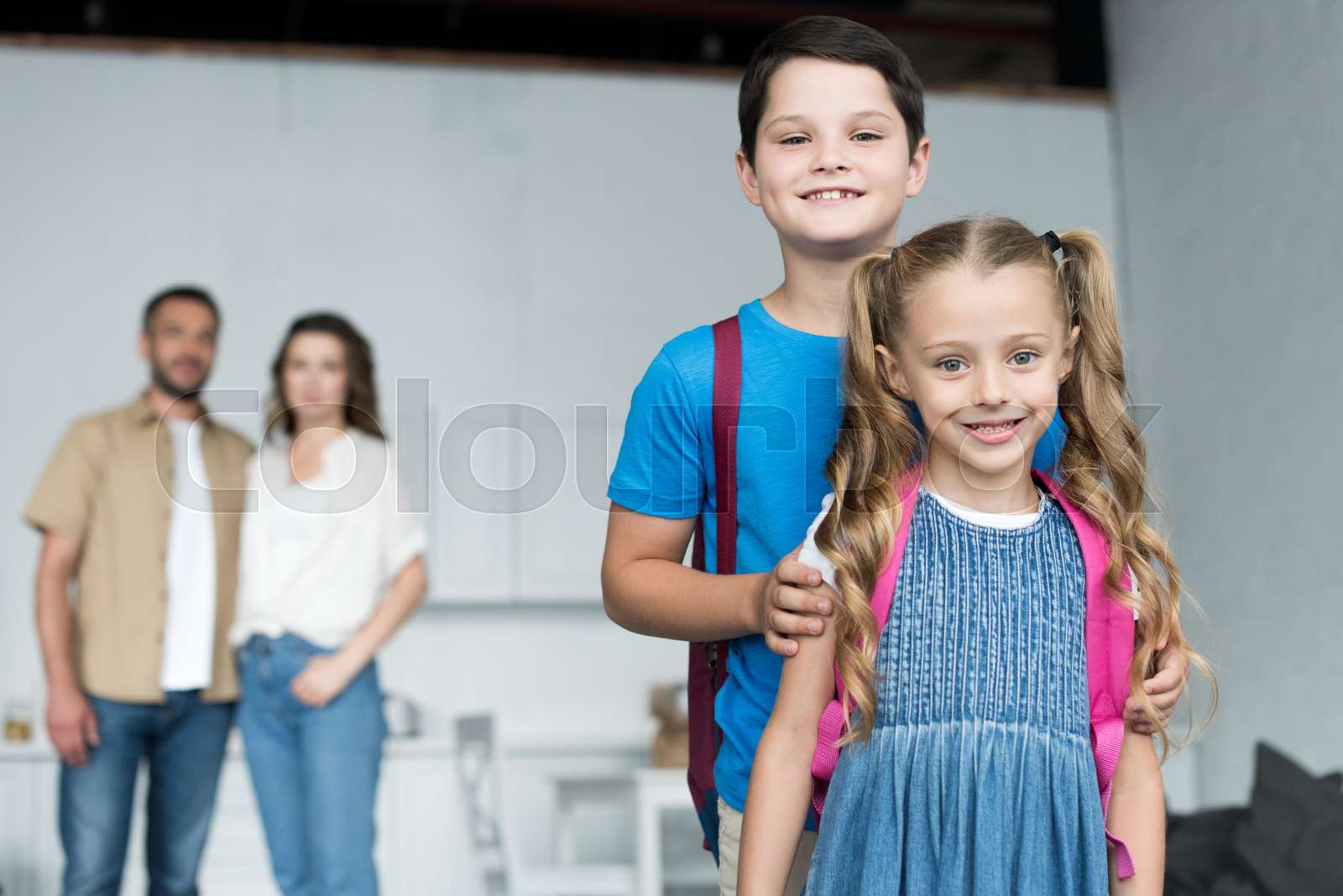 selective focus of smiling siblings with backpacks and parents behind ...