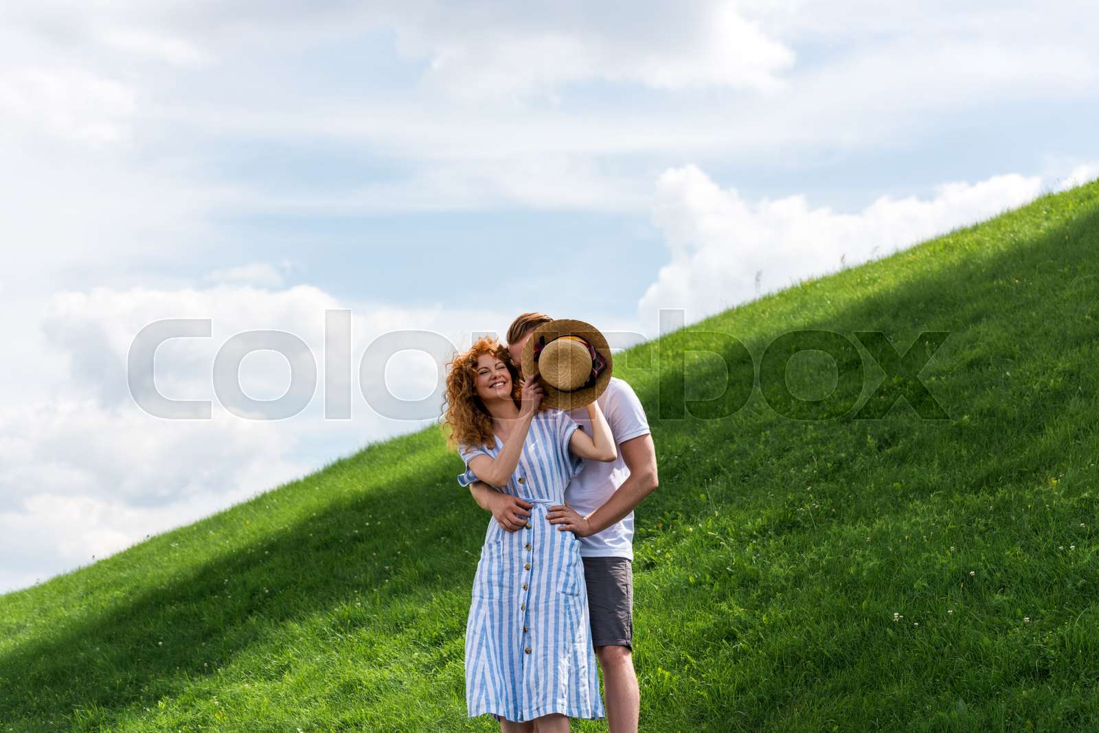 smiling redhead woman covering boyfriend face by straw hat on grassy ...
