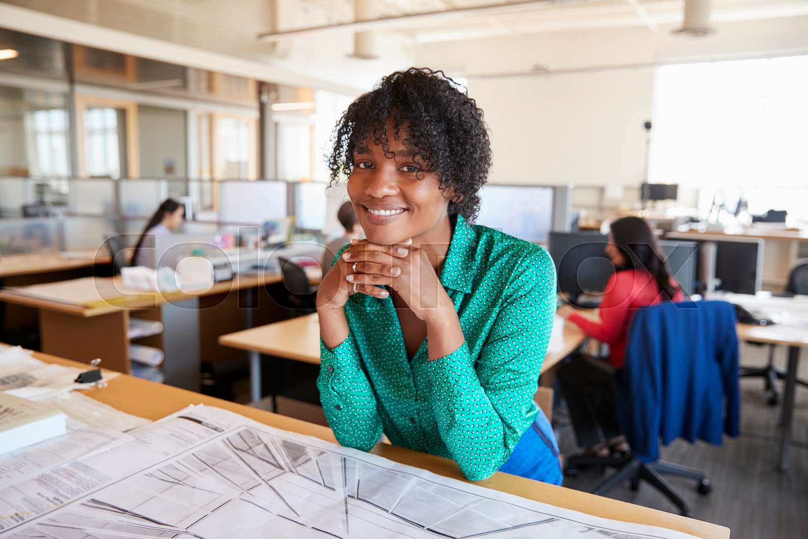 Black female architect leans on desk smiling in busy office | Stock image | Colourbox