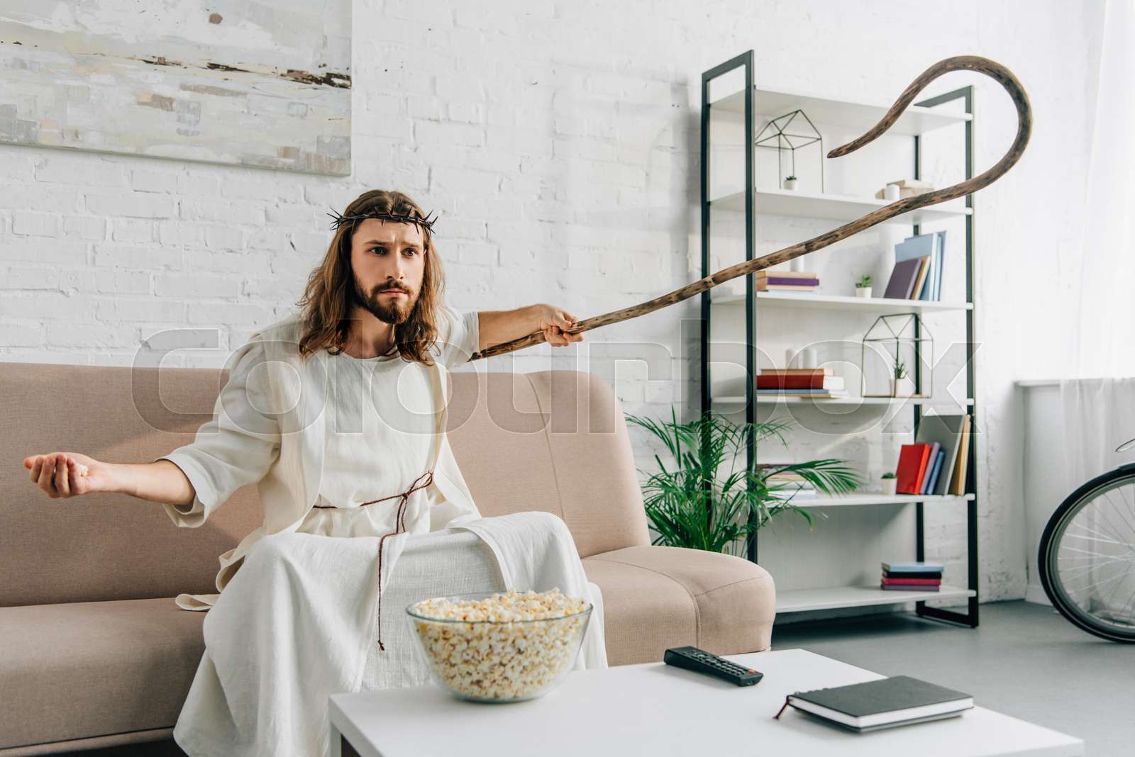 serious Jesus sitting on sofa with wooden staff and watching tv at home ...