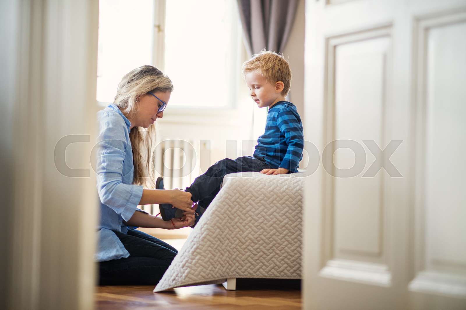 A young mother putting shoes on toddler son inside in a bedroom ...