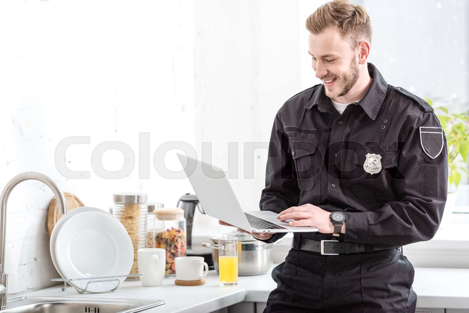 smiling police officer standing and using laptop at kitchen | Stock ...