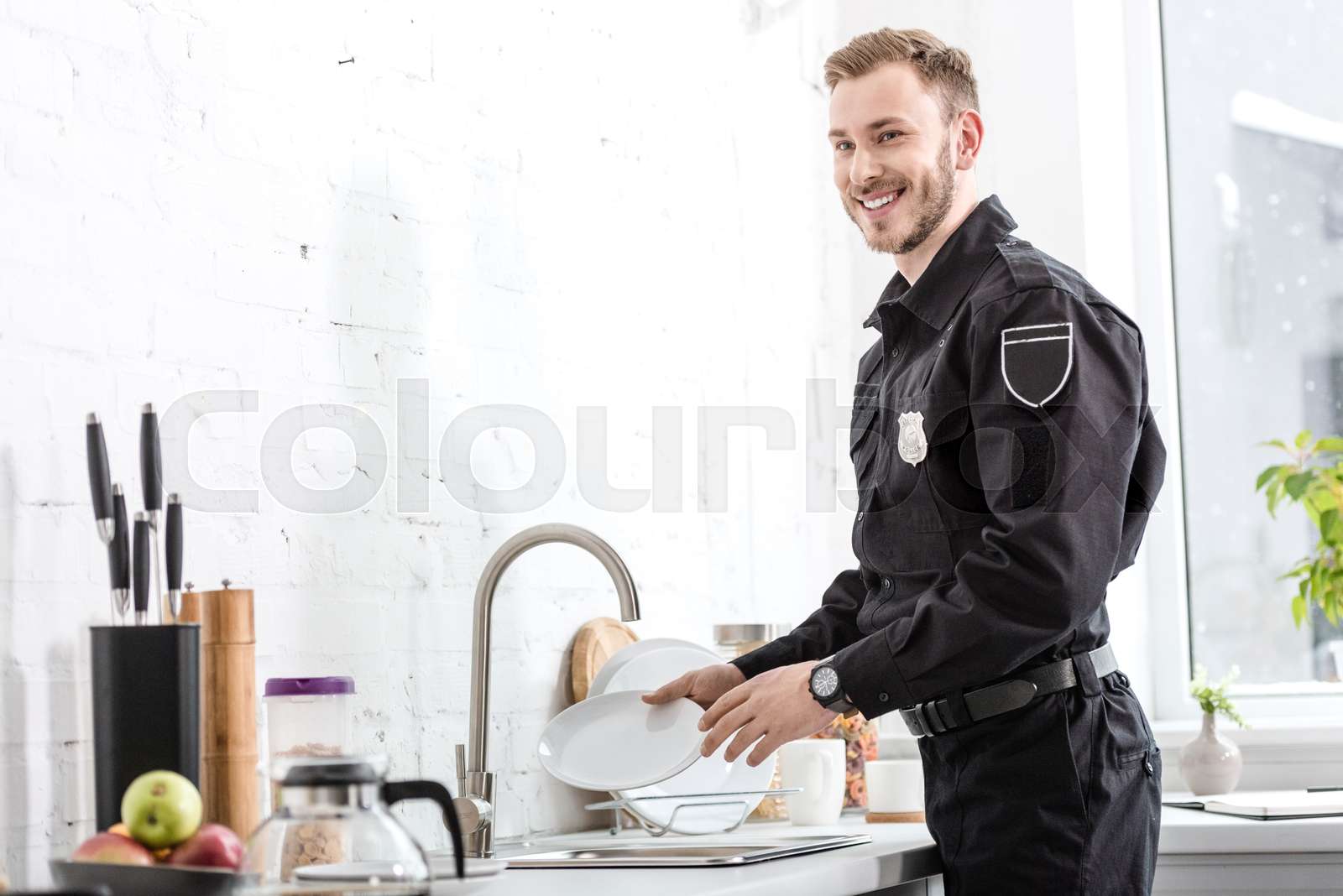 Handsome police officer smiling and washing plate at kitchen | Stock ...