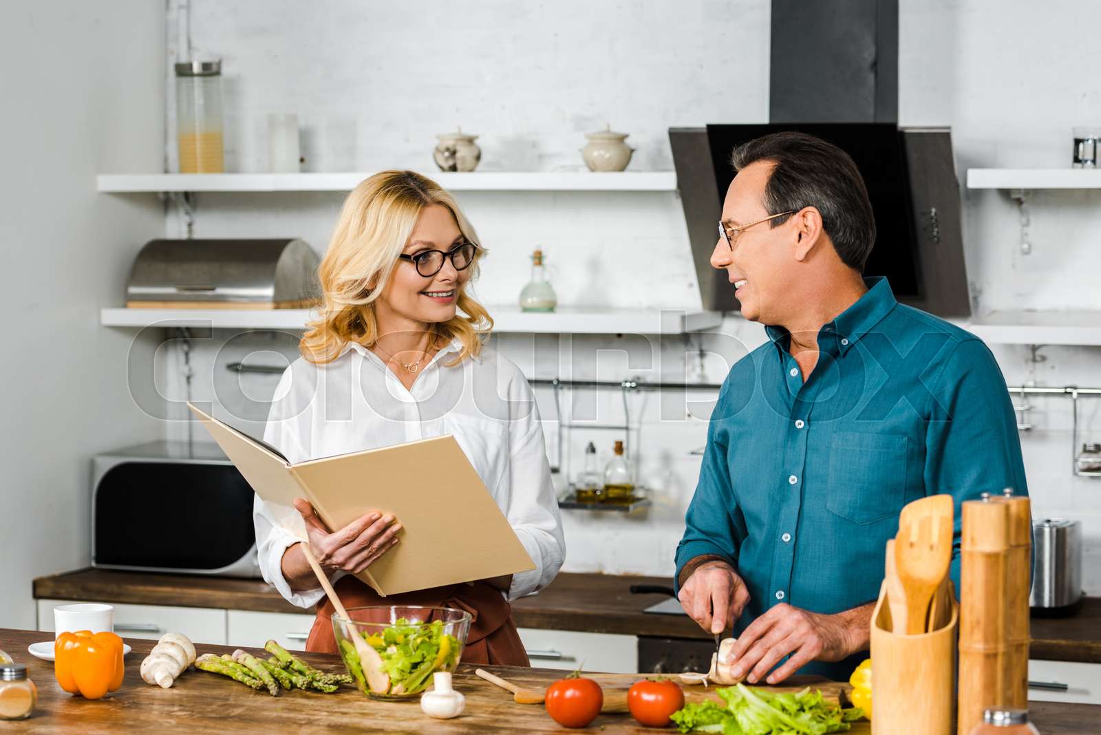 mature wife and husband cooking together with recipe book in kitchen ...