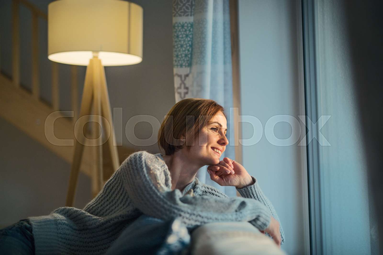 A young woman sitting indoors on a sofa at home, looking out of a ...