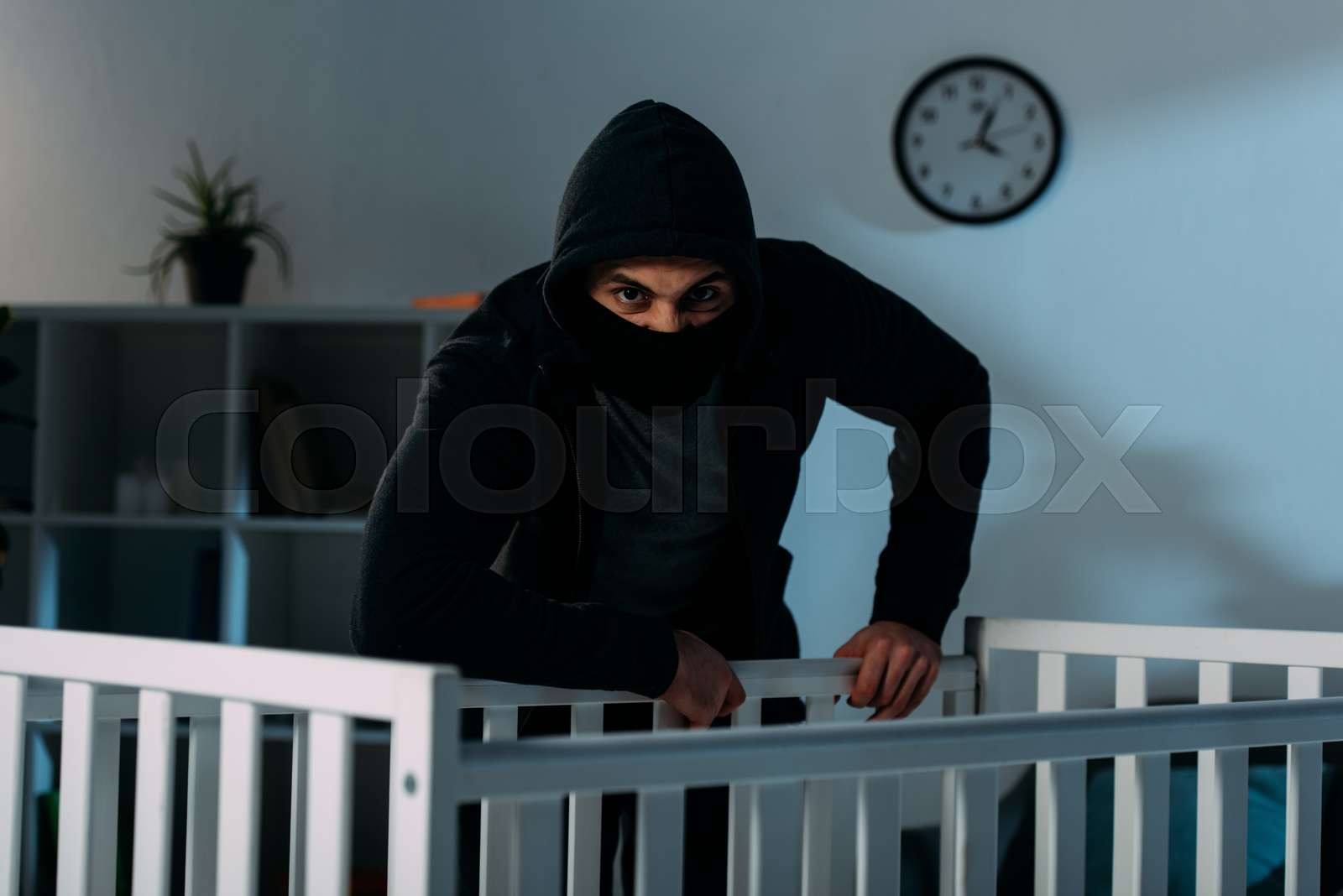 Angry kidnapper in black mask standing near crib and looking at camera ...