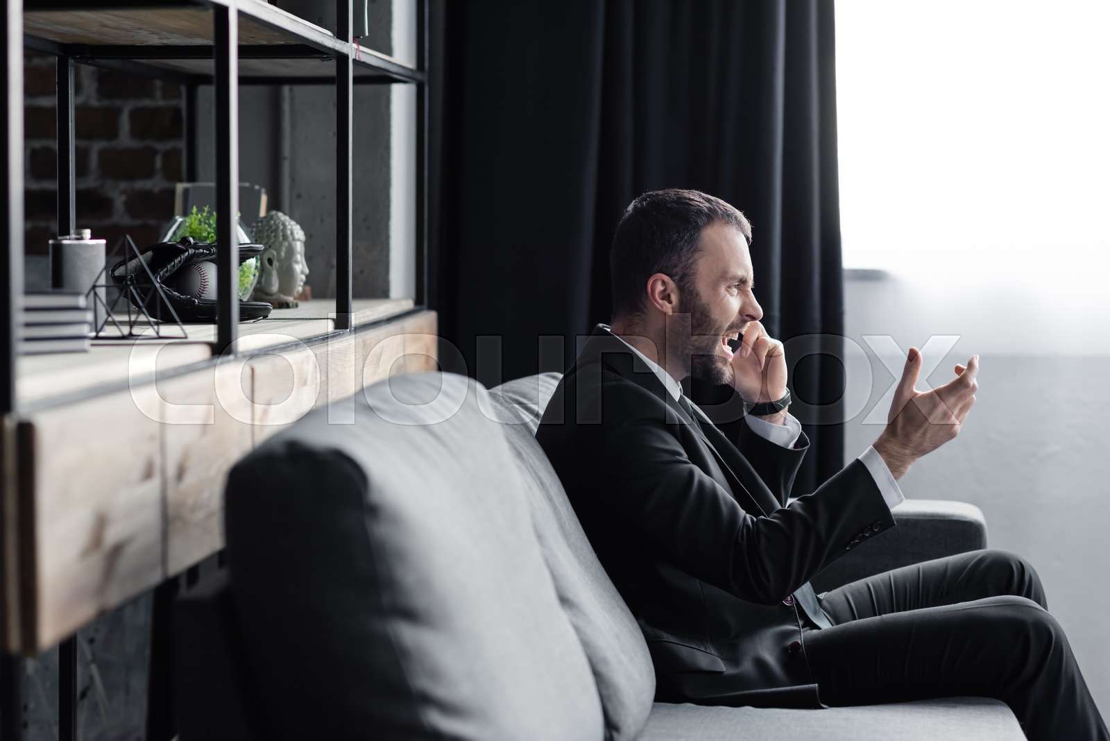 angry man sitting on sofa and quarreling while talking on smartphone in ...