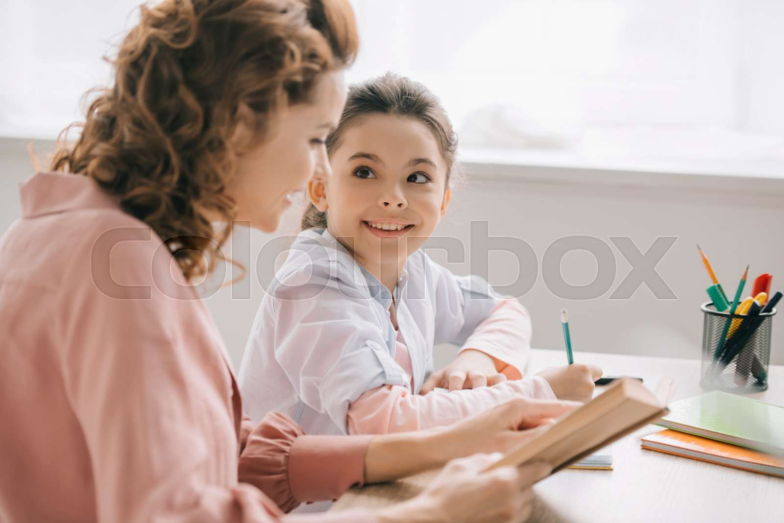 cheerful child looking at beautiful mother while doing homework ...