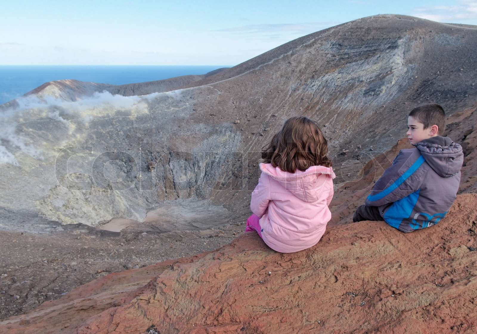 Girl and boy sitting on the rim of volcano crater of Vulcano island ...