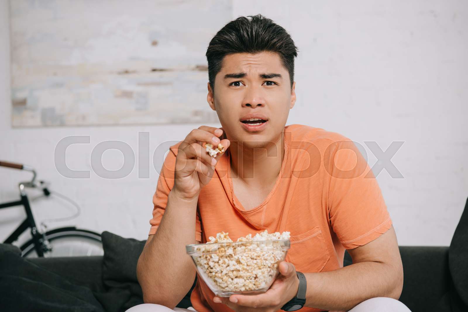 shocked asian man eating popcorn and watching tv while sitting on sofa at home | Stock image ...