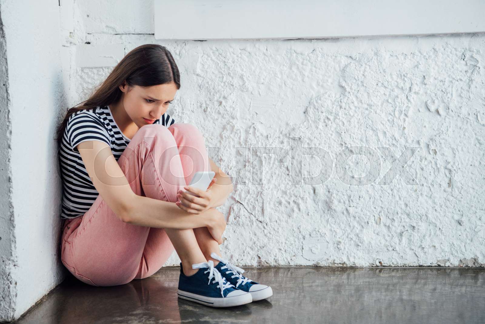 sad girl in pink pants sitting near wall and holding smartphone | Stock ...