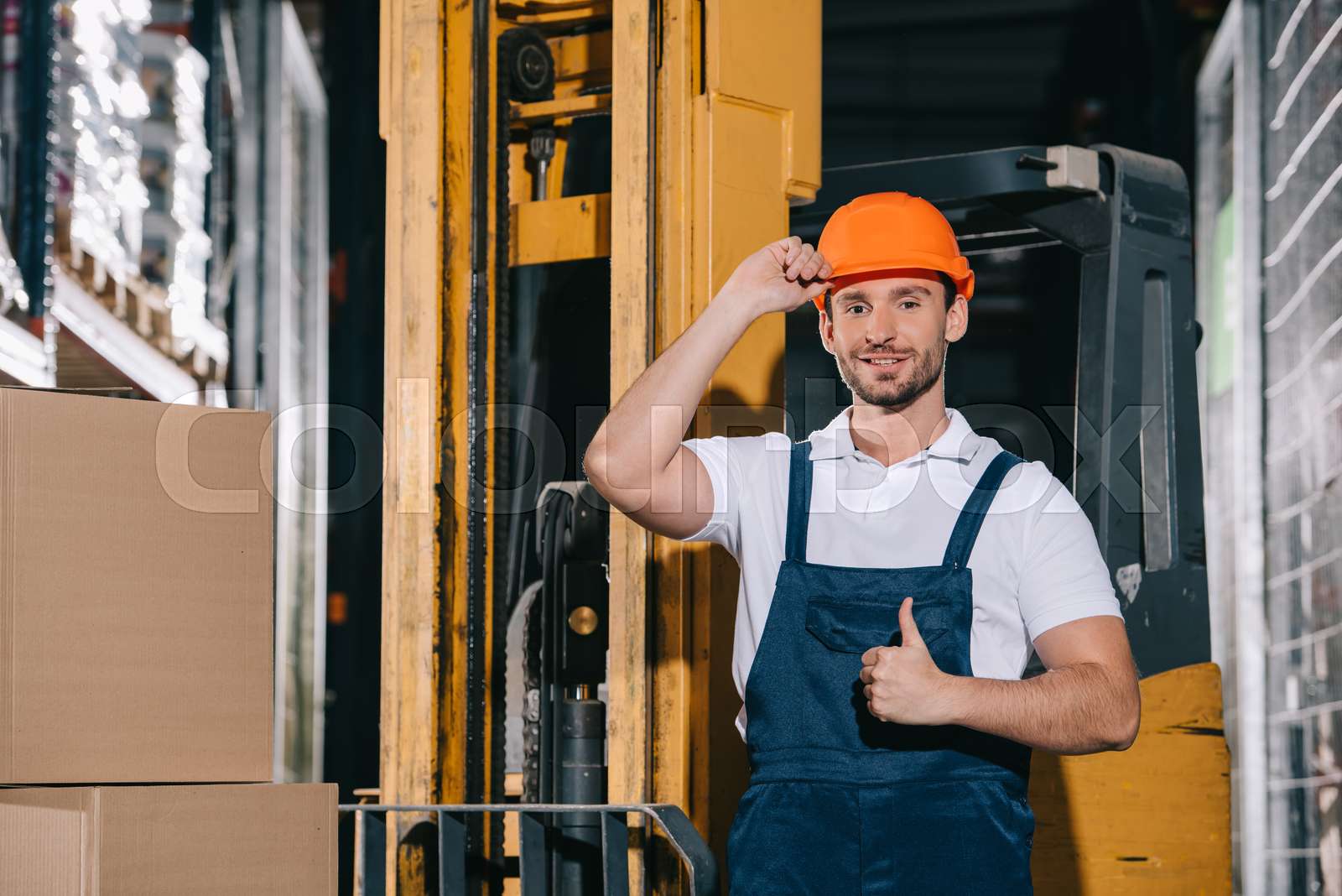 smiling warehouse worker touching helmet and showing thumb up while ...