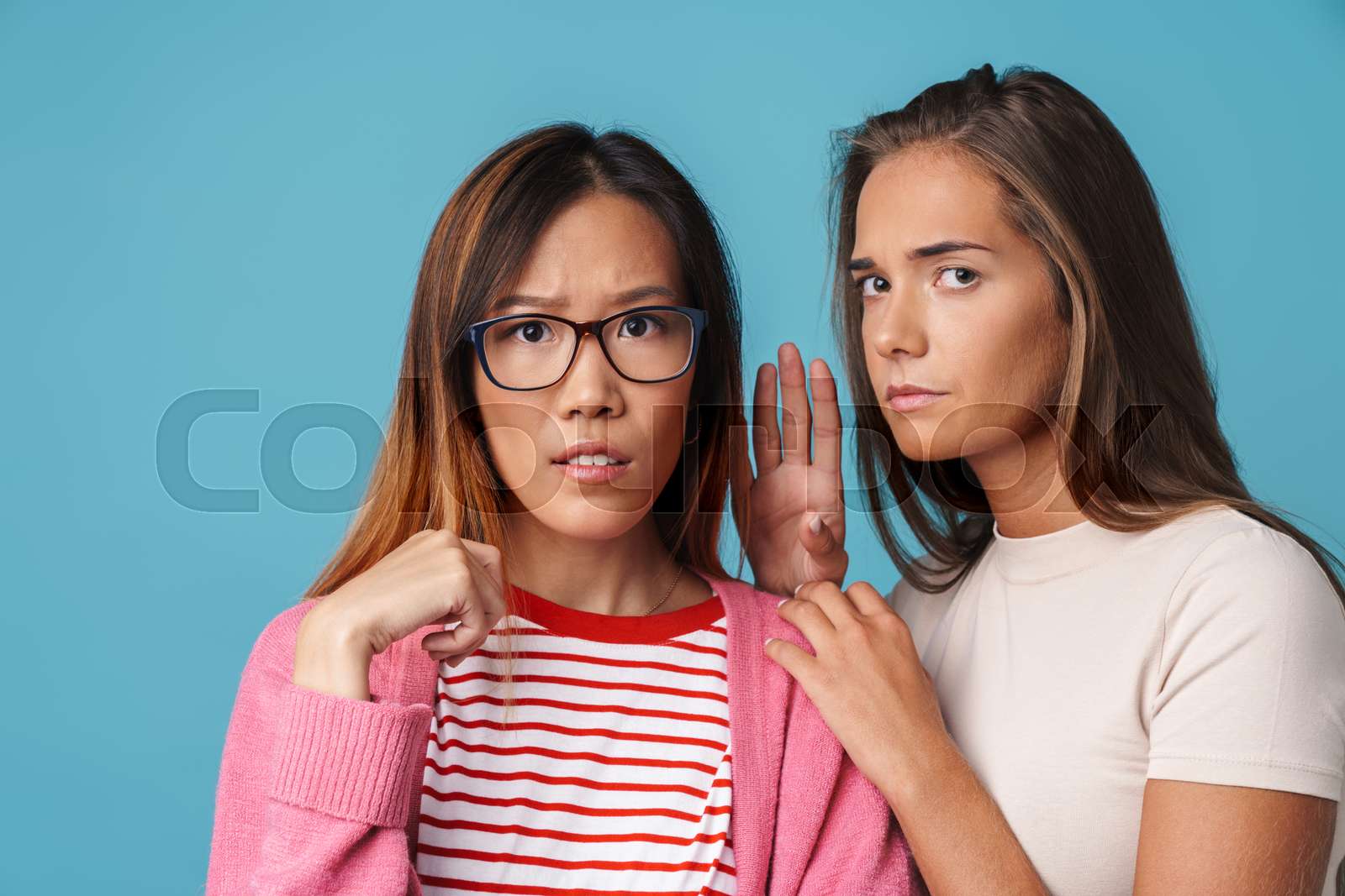 Portrait of two multinational confused women whispering secret in ear ...