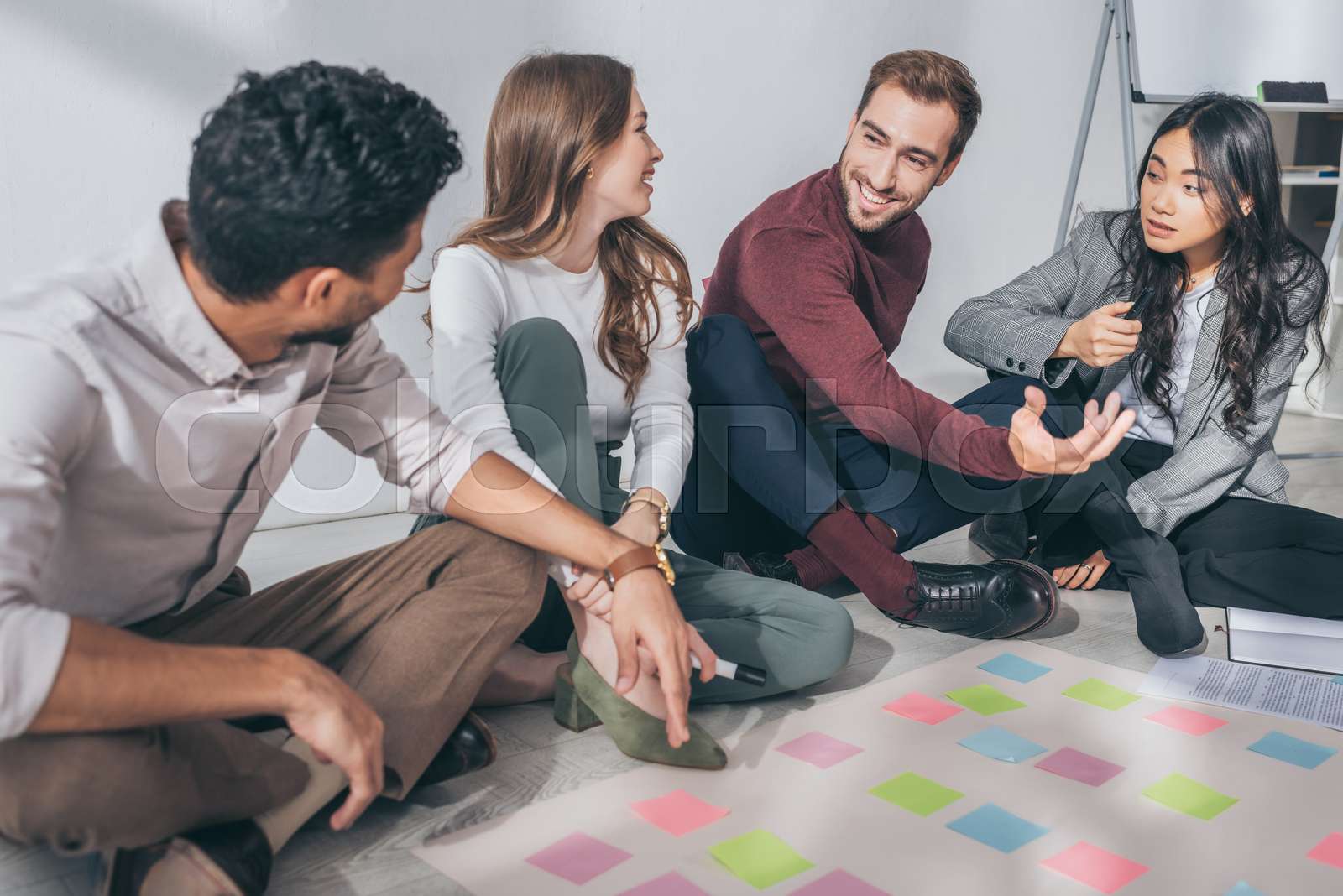 happy multicultural scrum masters sitting on floor near sticky notes ...