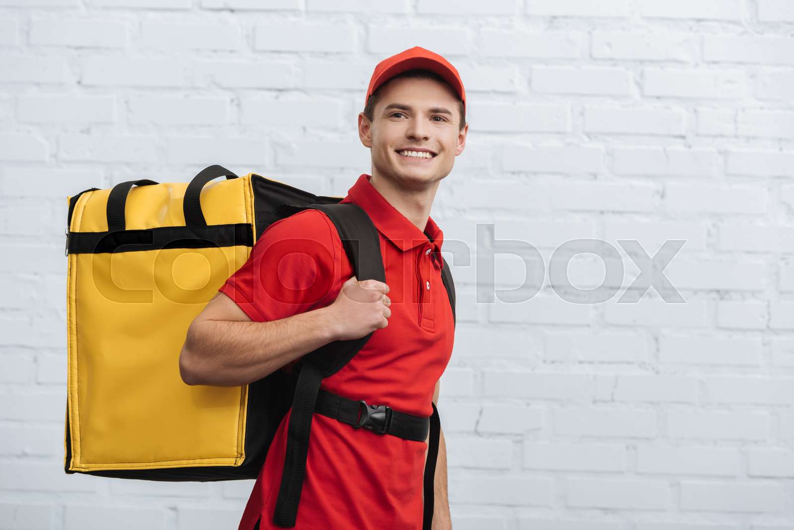 Smiling delivery man with thermo backpack looking away near brick wall ...