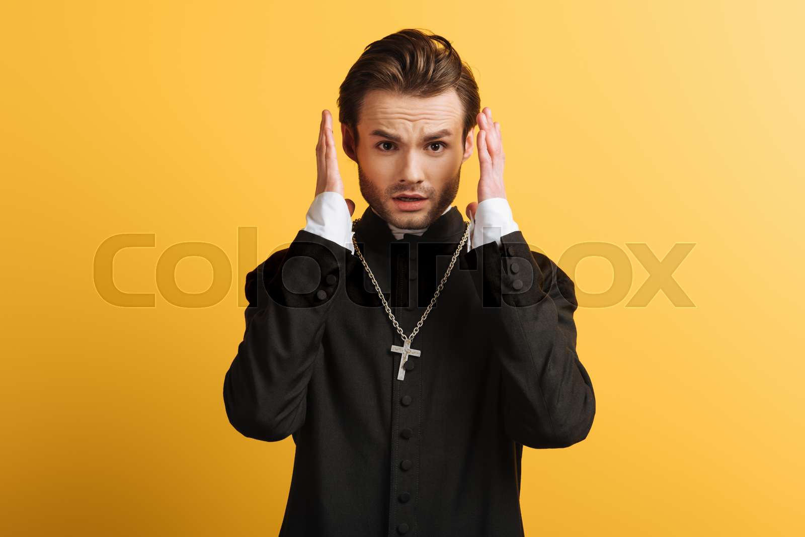 shocked catholic priest holding hands near head and looking at camera ...
