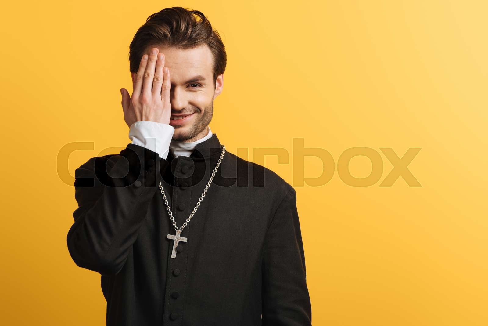 smiling catholic priest covering eye with hand while looking at camera ...
