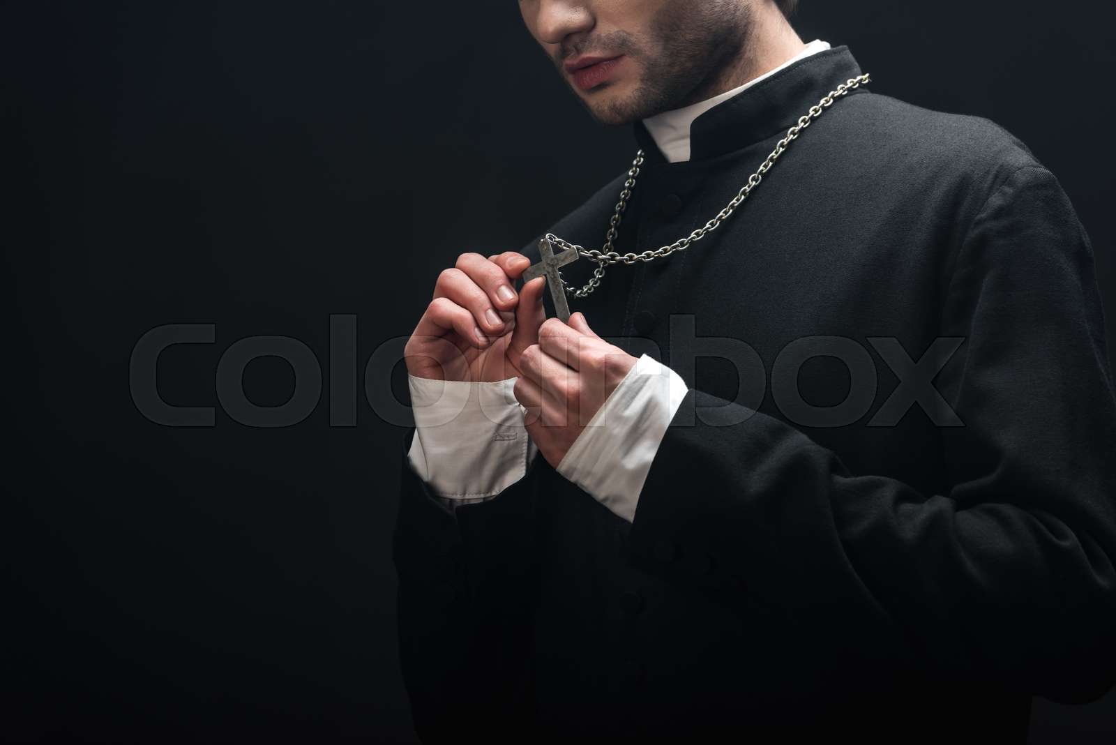 cropped view of catholic priest looking at silver cross on his necklace ...