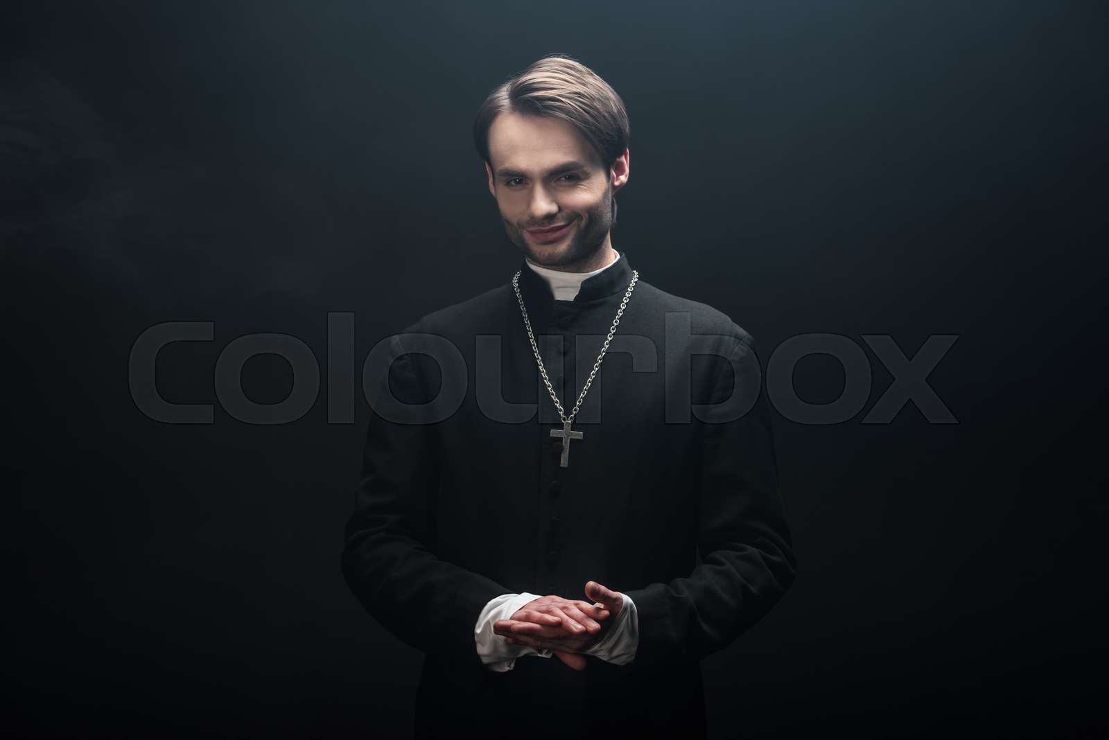 young smiling catholic priest standing with folded hands and looking at ...