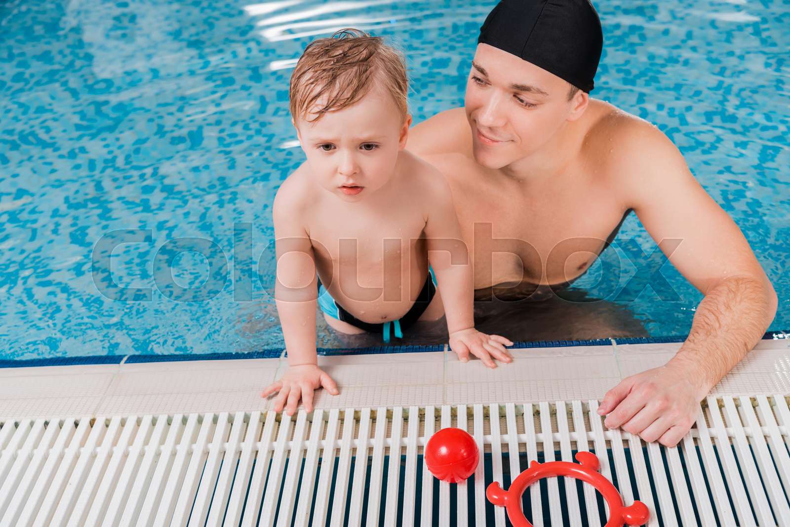 handsome swim coach in swimming cap looking at toddler boy in swimming ...