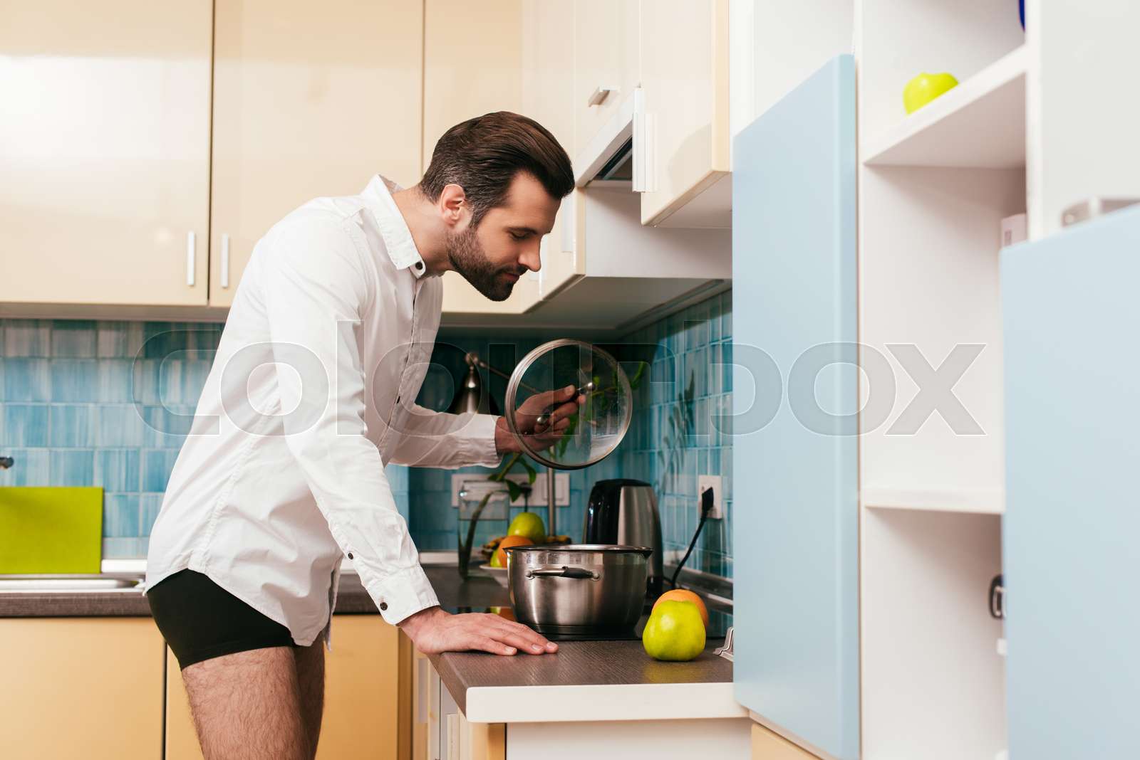 Side view of man in panties and shirt holding pan lid while cooking in ...