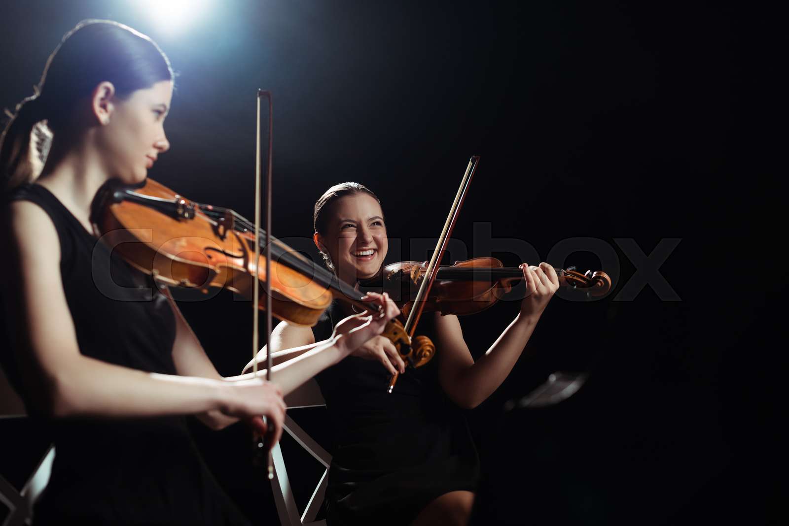 laughing female musicians playing on violins on dark stage with back ...