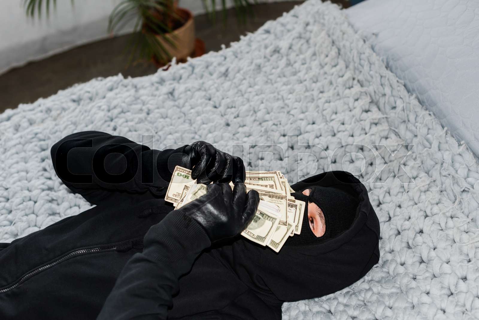 Robber in balaclava holding dollars near face while lying on bed ...