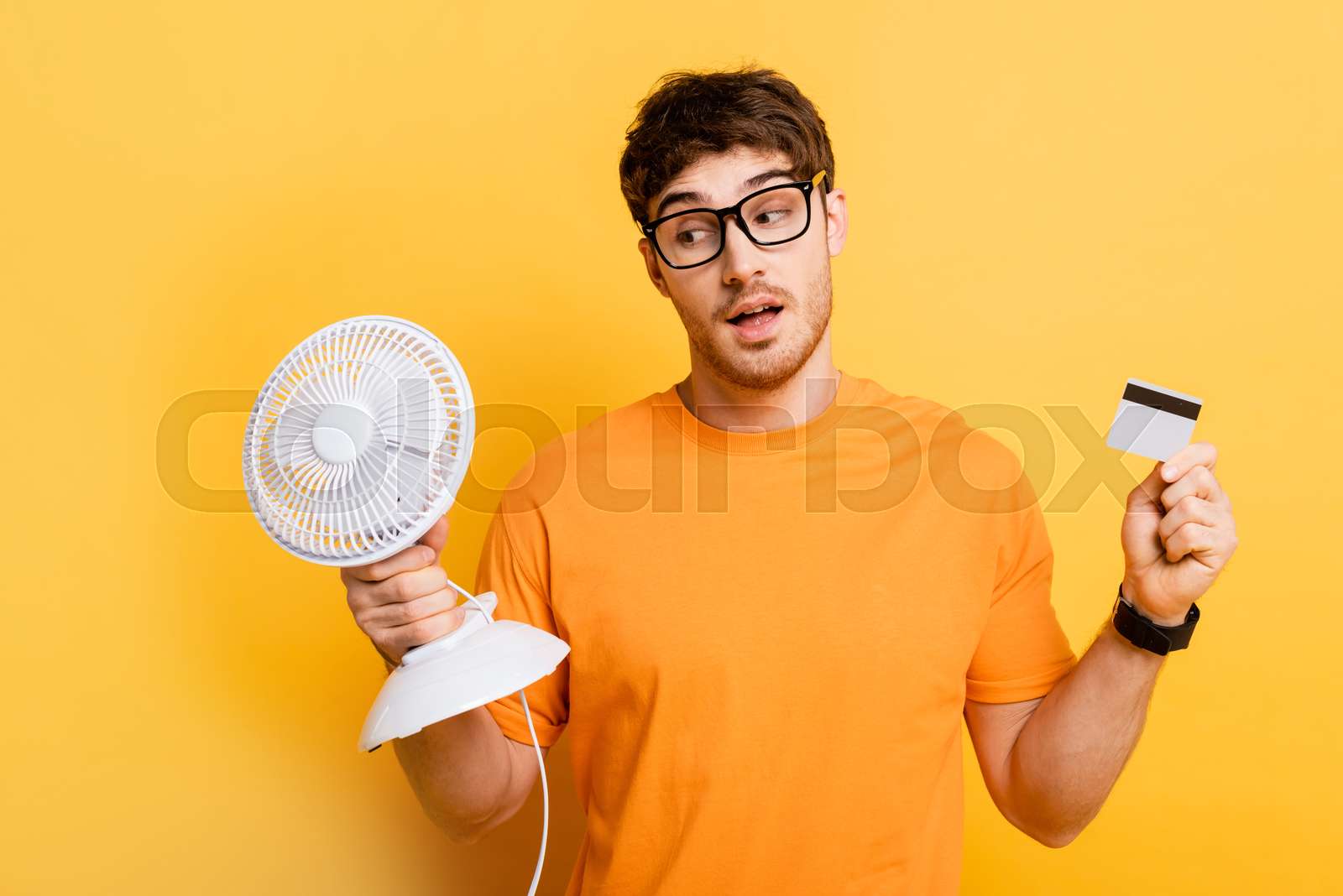 thoughtful young man holding electric fan and credit card on yellow ...