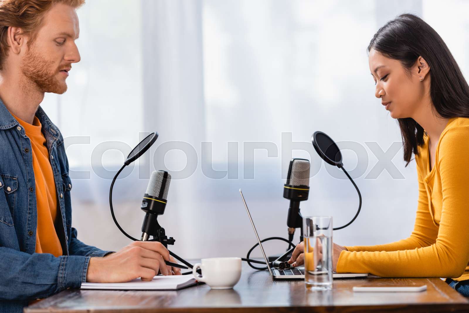 side view of asian broadcaster using laptop, and redhead man looking at ...