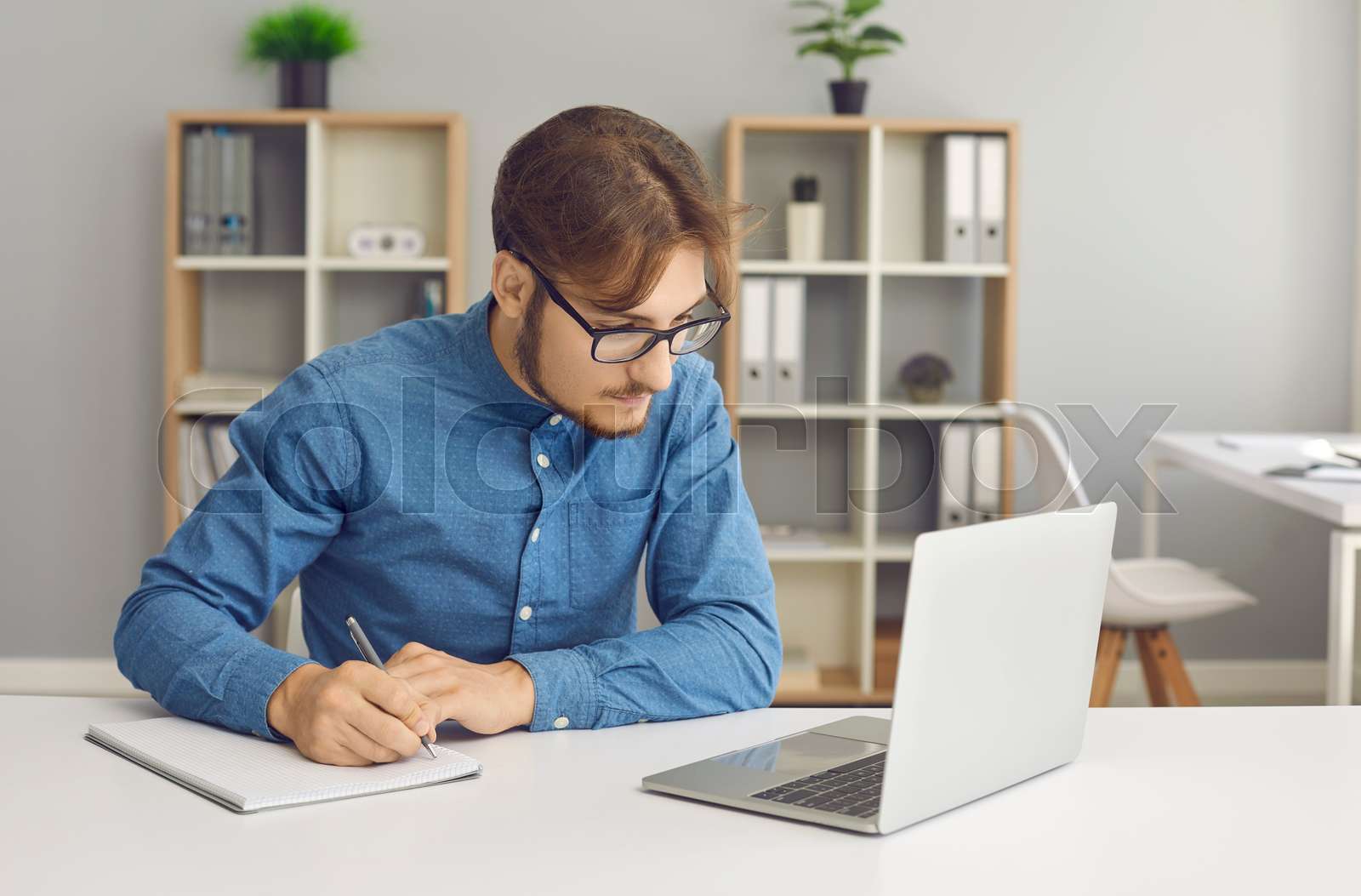 Concentrated young man with glasses makes notes while working with ...