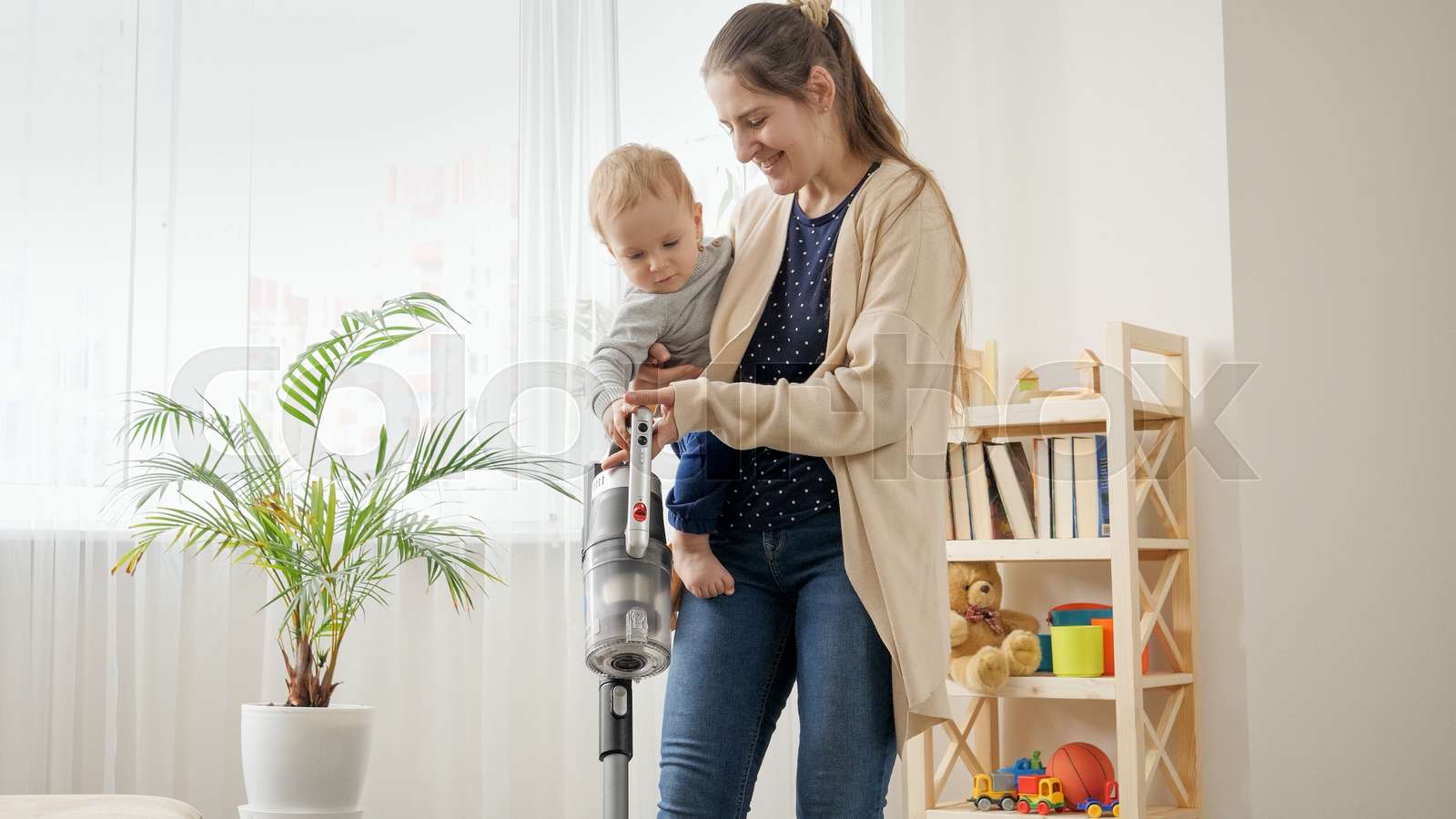 Young mother cleaning floor with vacuum cleaner while holding her ...