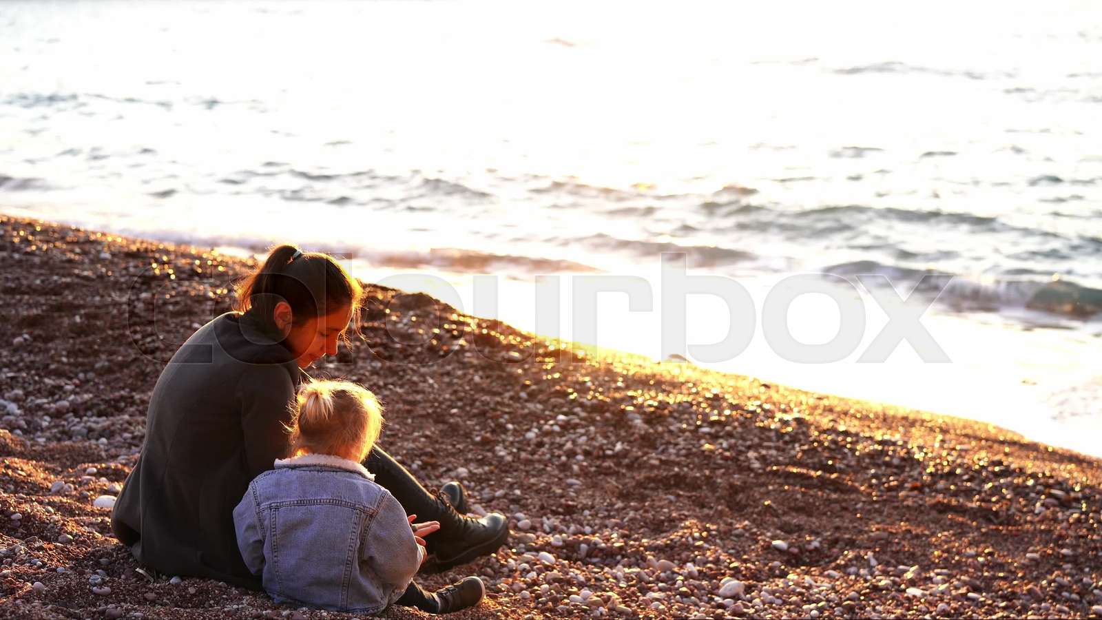 Mom and little daughter are sitting on a pebble beach and sorting ...