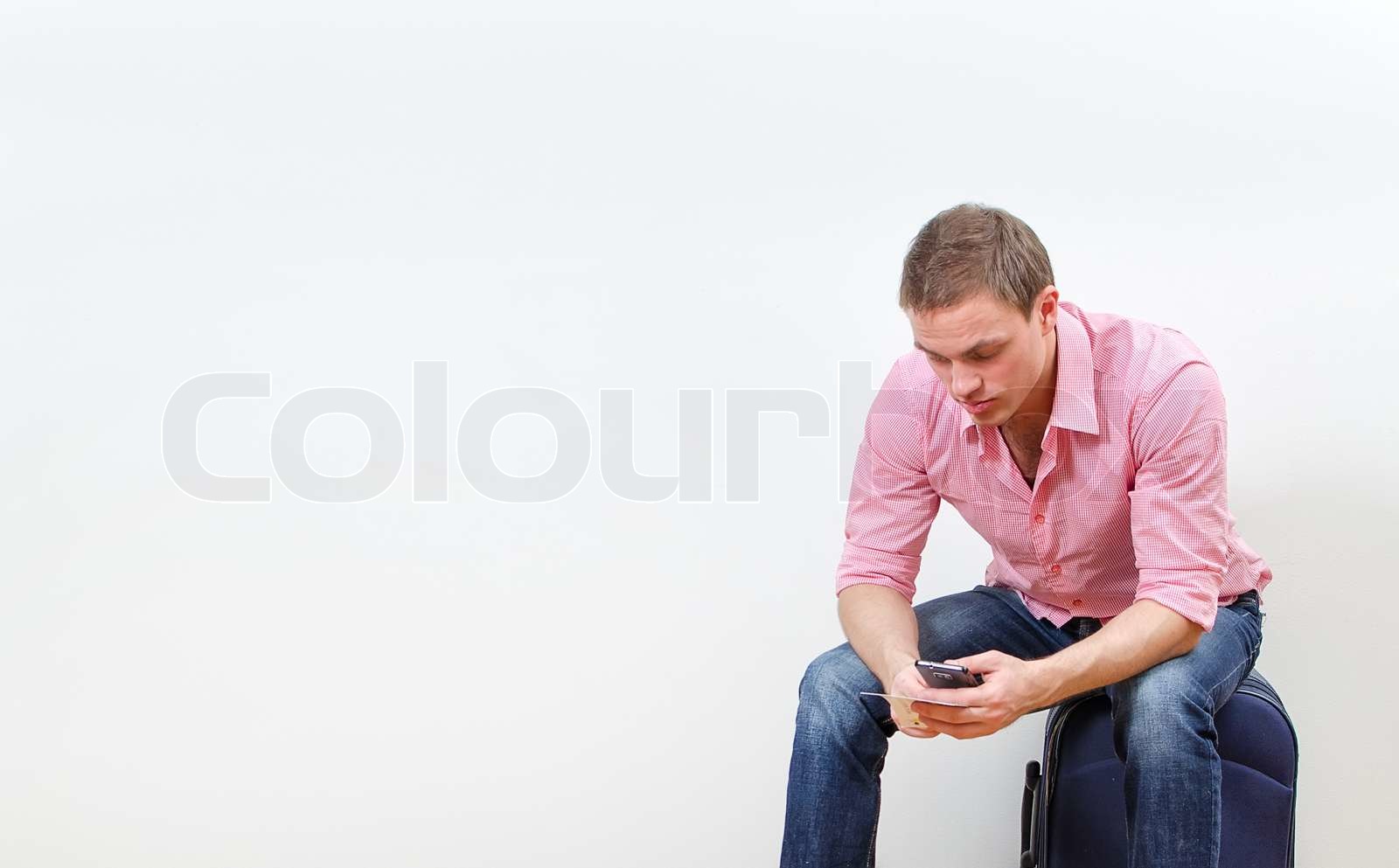 Junger Mann sitzt auf Koffer und mit seinem Handy | Stock Bild | Colourbox