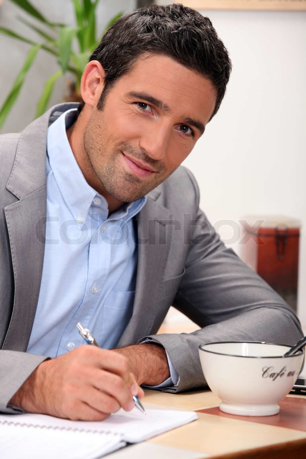 charming 30 years old man dressed in a grey suit and writing in a cosy ...